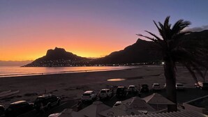 Um estacionamento à beira-mar com carros e uma palmeira, com vista para um pôr do sol e montanhas ao fundo.