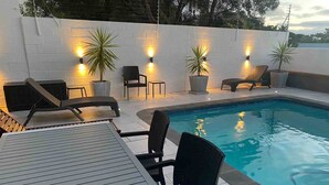 Uma área à beira da piscina com mesa e cadeiras, cercada por plantas em vasos e uma parede de tijolos brancos.