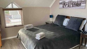 Quarto com cama grande, janela com persianas, mesa de cabeceira, luminária e quadro na parede.