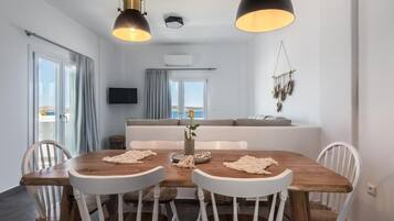 Uma sala de jantar com mesa de madeira e cadeiras brancas, TV montada na parede e vista para o exterior através de grandes janelas.
