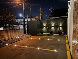 Área de estacionamento fechada à noite, com plantas em vasos iluminadas e um caminho bem definido marcado por linhas amarelas.