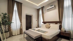 Quarto de hotel moderno com uma cama grande, um banquinho, uma mesinha de cabeceira e um vaso com planta.