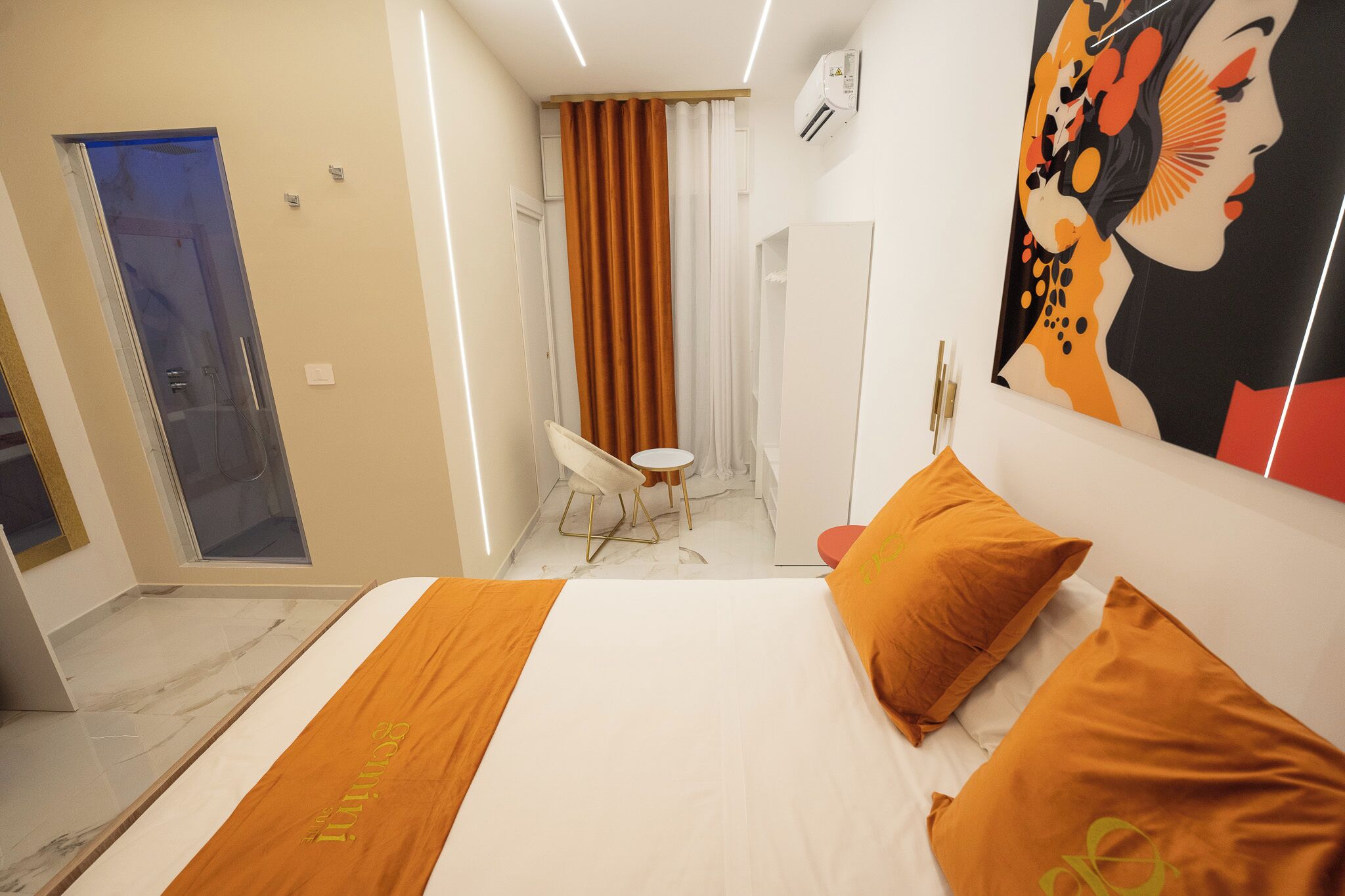 Quarto de hotel com cama, manta laranja, cadeira, mesinha e um quadro na parede.