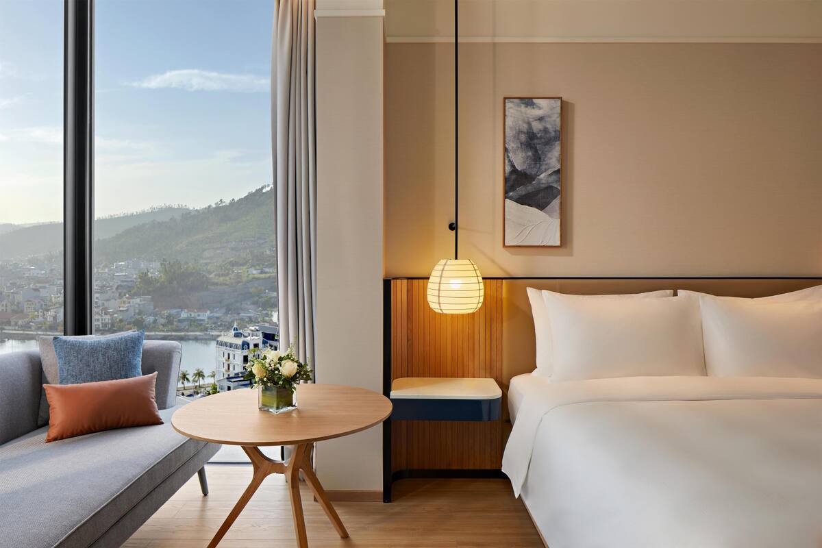 Quarto de hotel com uma janela grande, uma cama, um sofá, uma mesa pequena com um vaso de flores e uma luminária fixada na parede.
