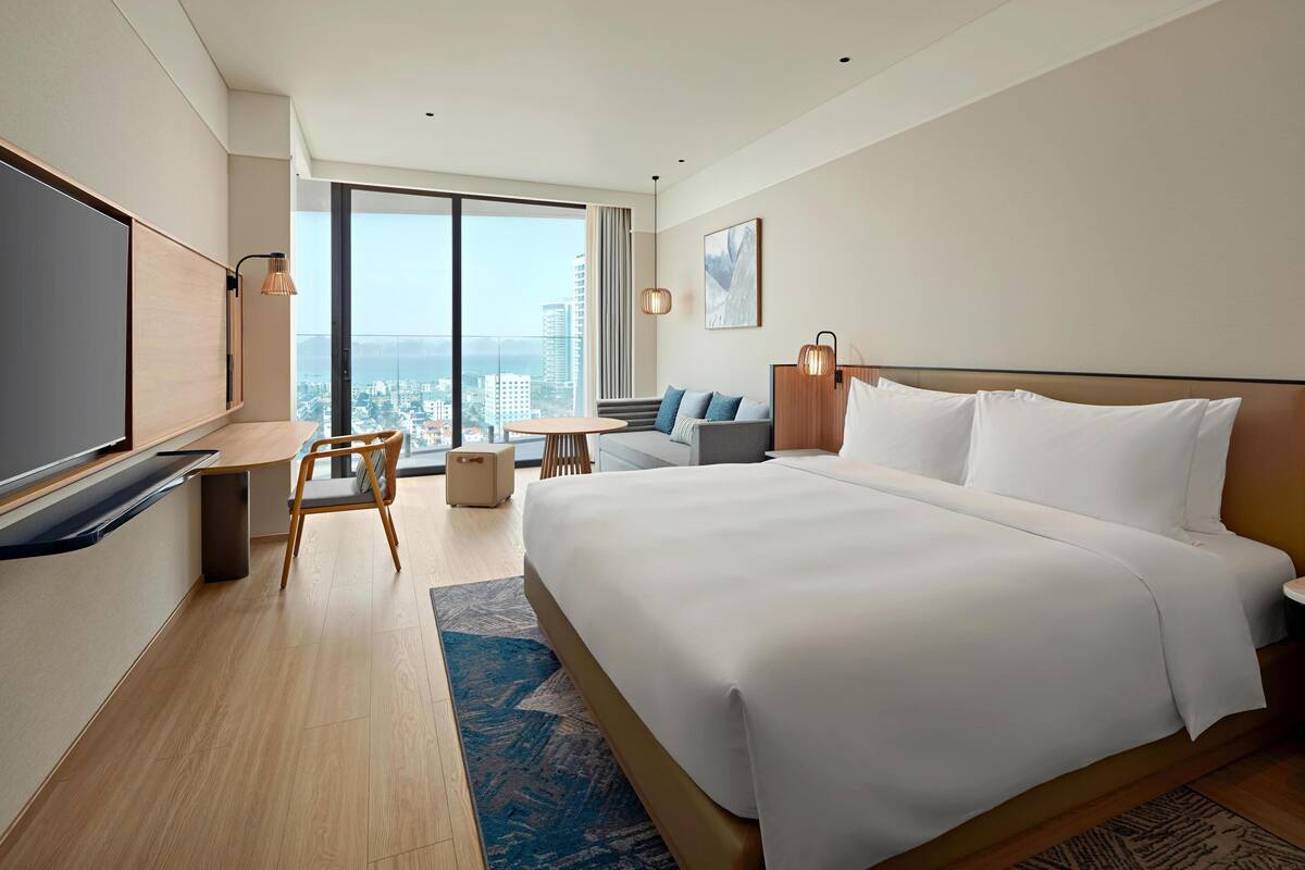 Quarto de hotel moderno com uma cama grande, escrivaninha, cadeira, televisão e vista para a cidade.