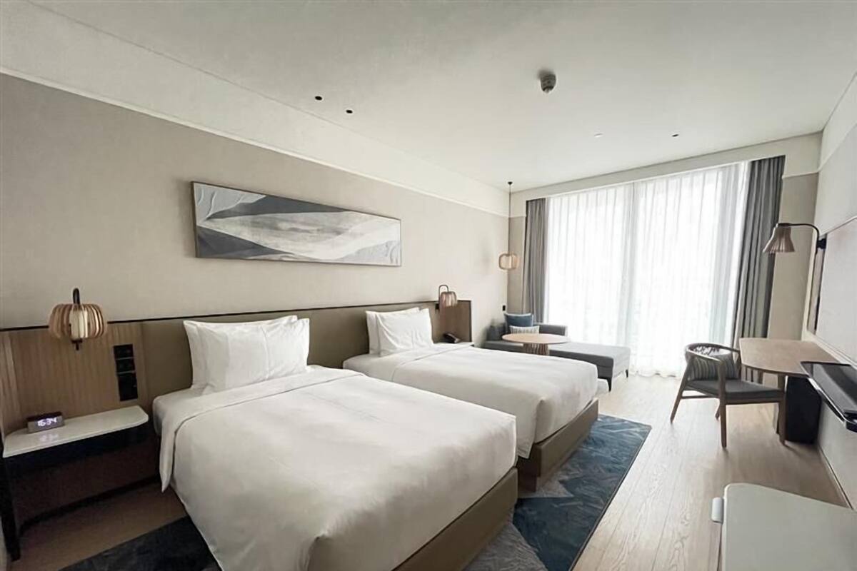 Quarto de hotel com duas camas, uma escrivaninha, uma cadeira, uma televisão e uma janela grande com cortinas.