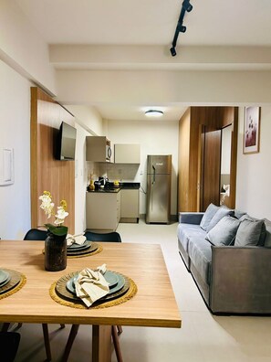 Cozinha moderna com geladeira, fogão e armários. Há uma área de jantar com mesa posta para dois, um sofá e uma TV fixada na parede.