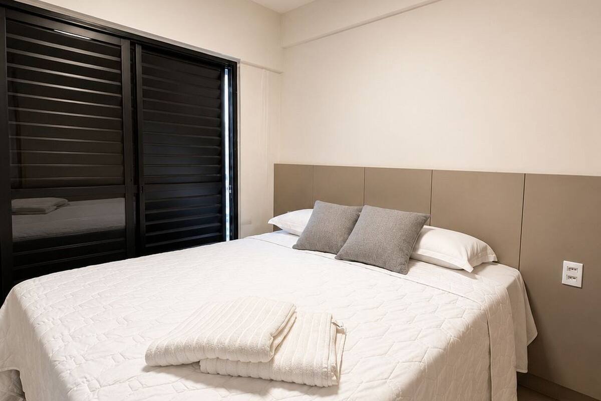 Um quarto com uma cama grande, cabeceira, janela com persianas e luminária no teto.