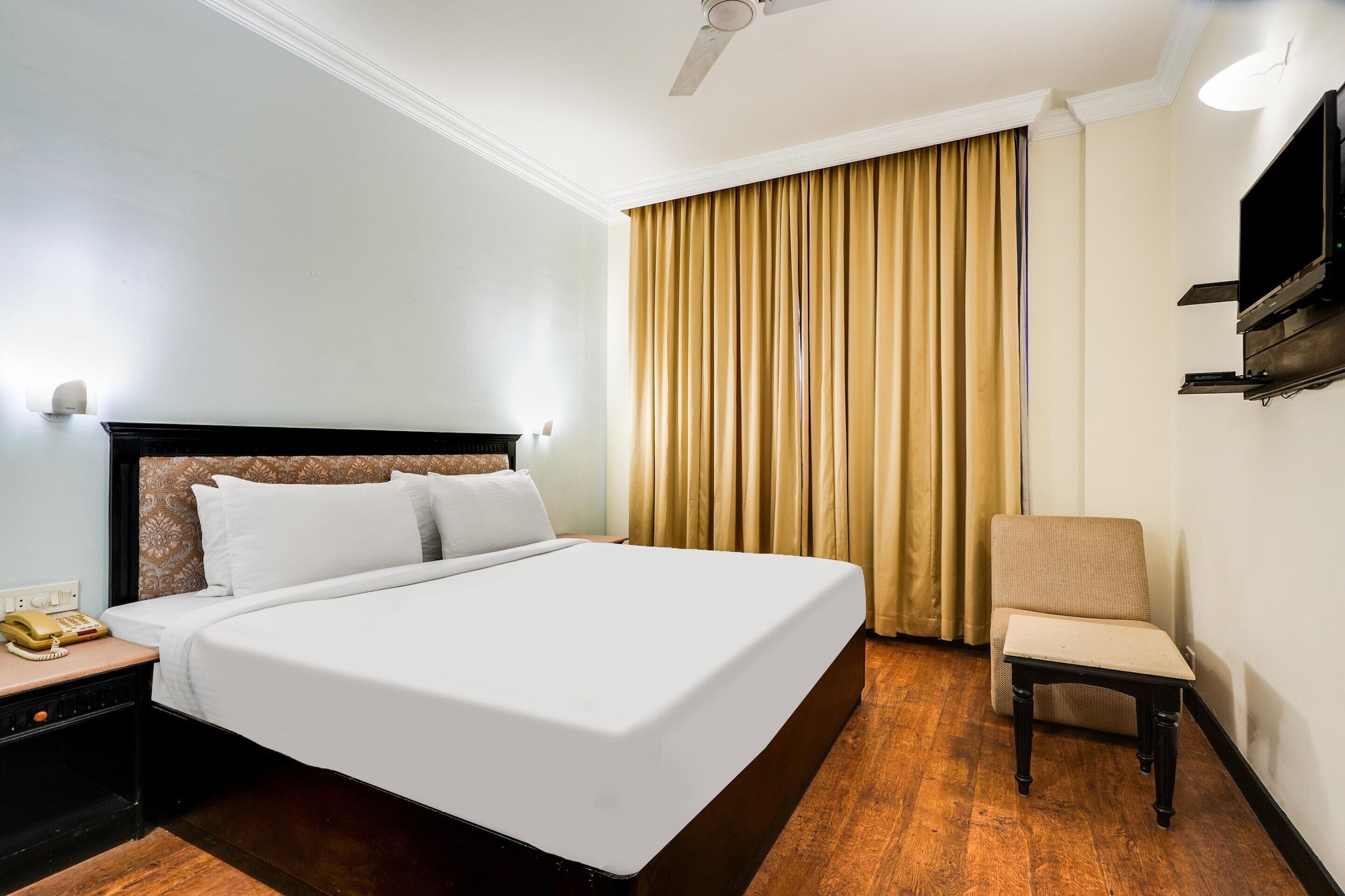 Quarto de hotel com uma cama grande, uma cadeira, escrivaninha com telefone e televisão na parede.