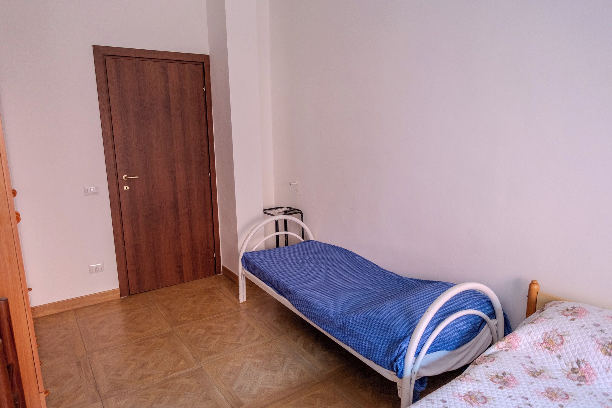 Quarto pequeno com uma cama de solteiro, porta de madeira, estrutura de cama de metal branca e colchão azul.