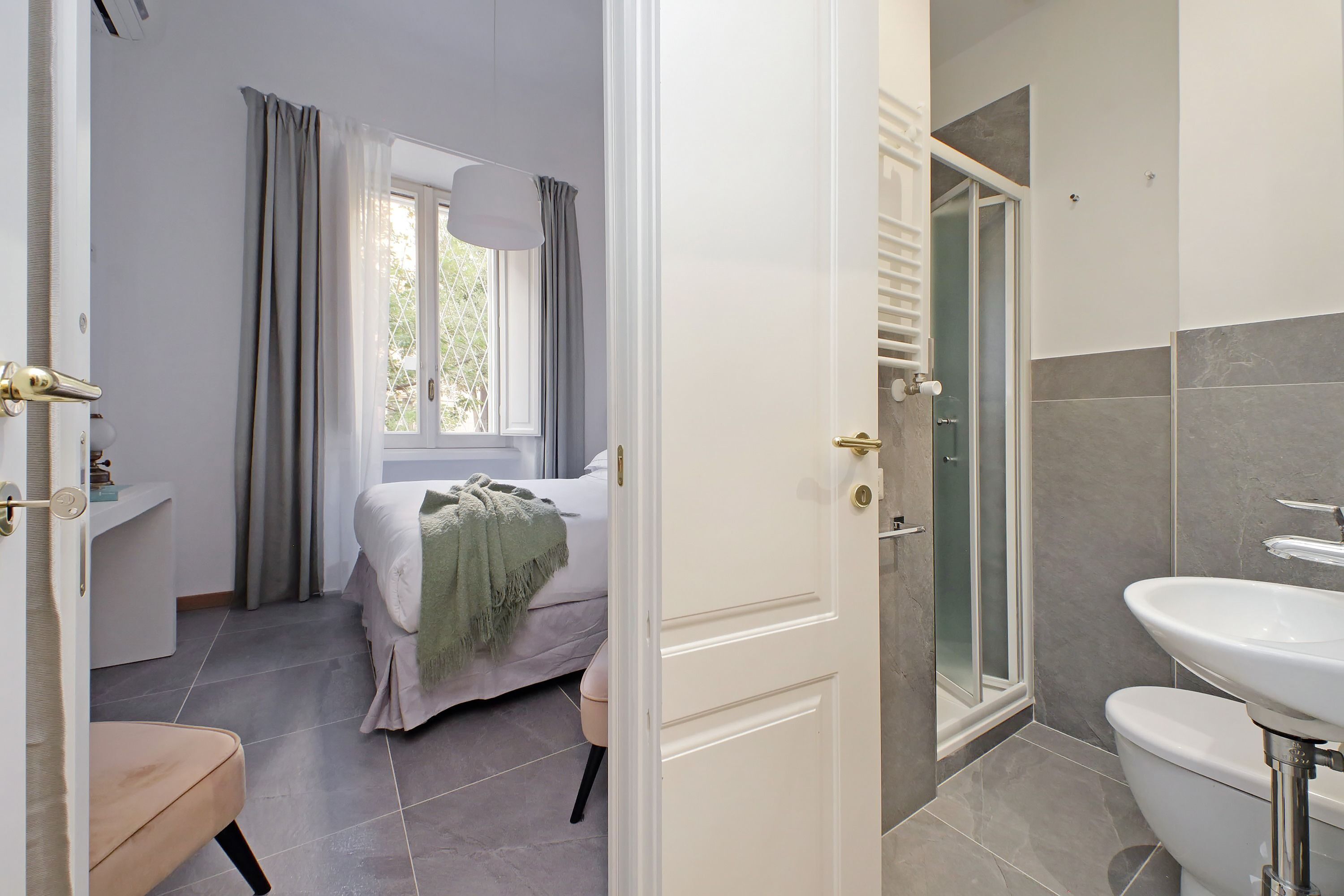 Quarto com cama, mesa de cabeceira, janela com cortinas e banheiro com pia e chuveiro.