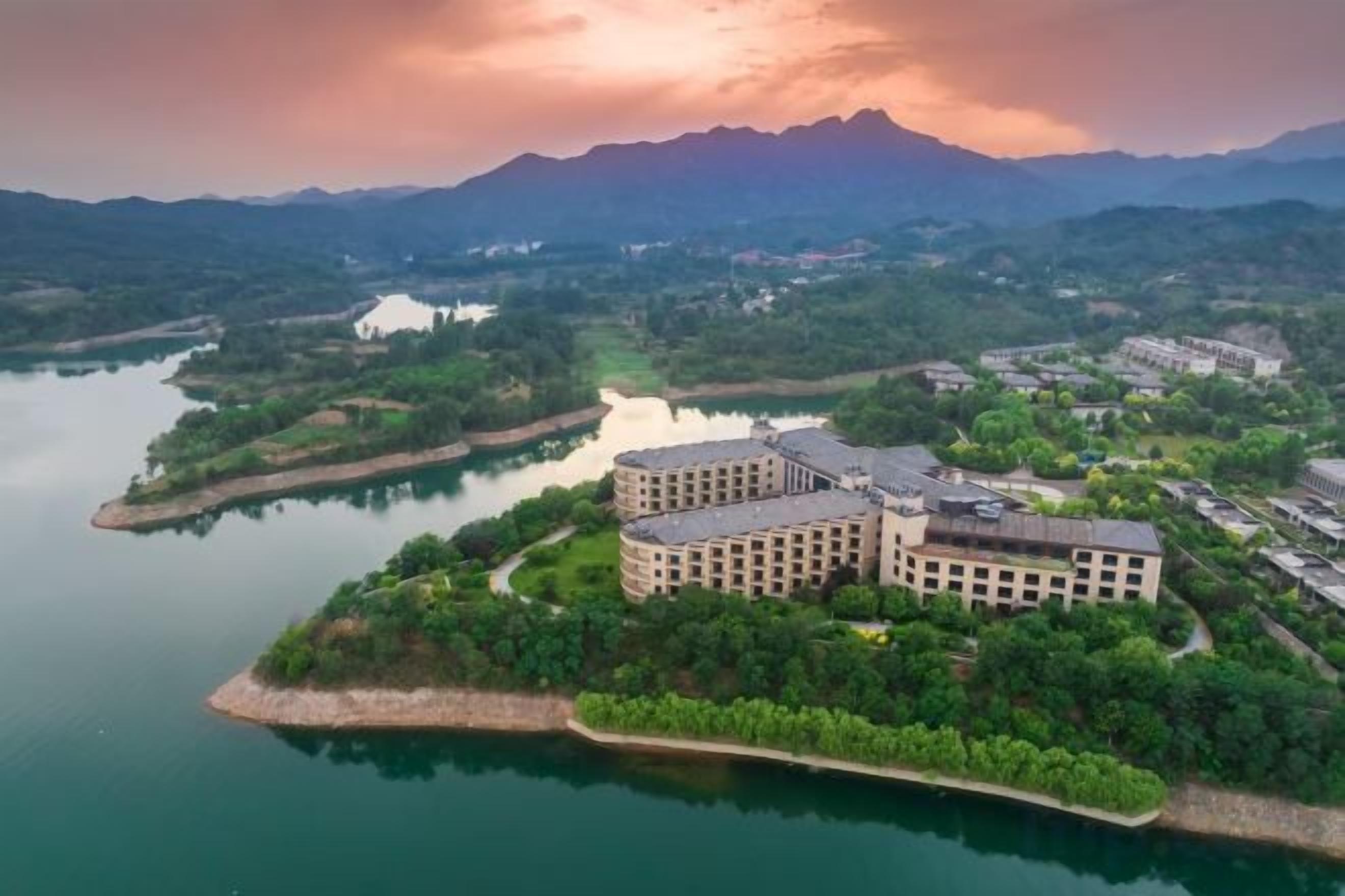 Um hotel com vários edifícios, cercado por água e vegetação, com montanhas ao fundo.