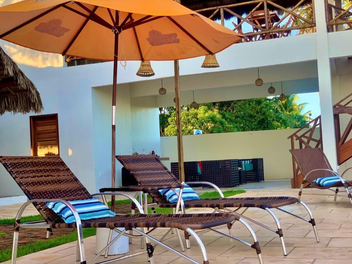 Uma área de pátio com cadeiras de lounge e guarda-sol, uma estrutura com telhado de palha e vista para o verde.