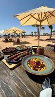 Um deck de madeira com mesas para refeições ao ar livre, guarda-sóis e diversos pratos, incluindo carnes grelhadas e uma salada.