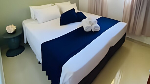 Uma cama bem arrumada com roupa de cama branca e azul marinho, uma pequena mesa de cabeceira com uma flor e uma janela com cortinas.