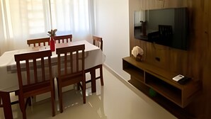 Uma área de refeições com mesa e cadeiras, uma televisão fixada em uma parede de madeira e uma janela com cortinas.