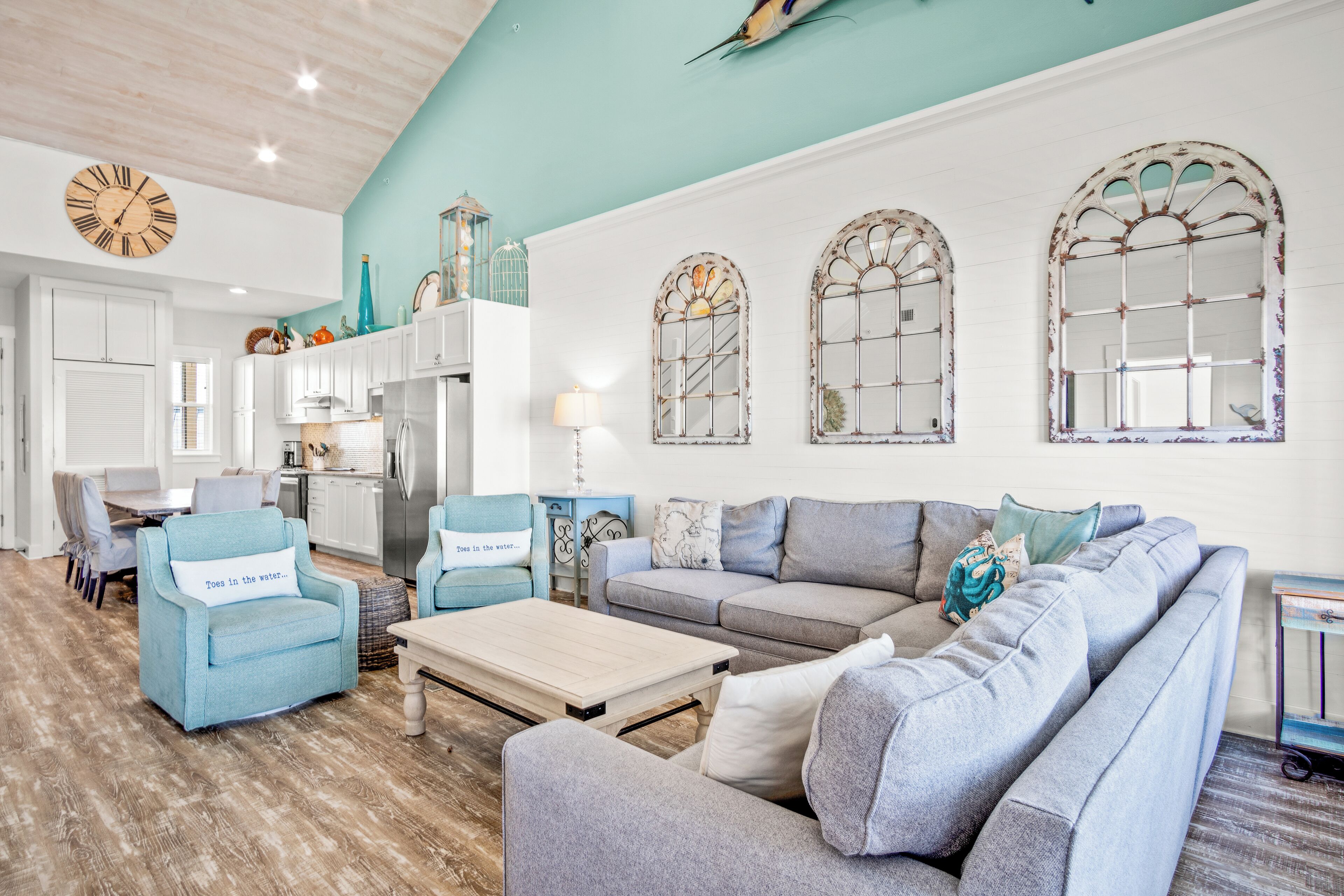 Uma sala de estar ampla, com um sofá cinza, uma poltrona azul e uma mesa de centro de madeira. Há uma área de cozinha com armários brancos e bancada azul.