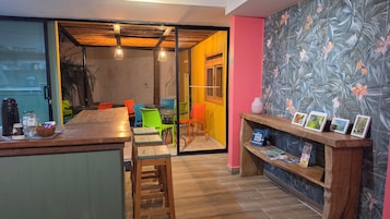 Quarto com mesa de madeira, cadeiras coloridas, estante com fotos emolduradas e papel de parede tropical.