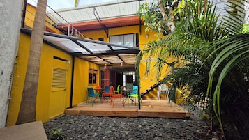 Um edifício amarelo com um terraço coberto, mesas ao ar livre e uma escadaria que leva a um andar superior.