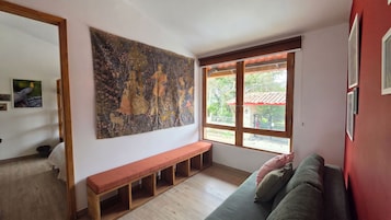 Quarto com um banco de madeira, um sofá verde, uma tapeçaria grande e uma janela com vista para vegetação e um edifício.