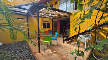 Uma área de pátio com deck de madeira, cadeiras coloridas e um edifício amarelo com porta de vidro.