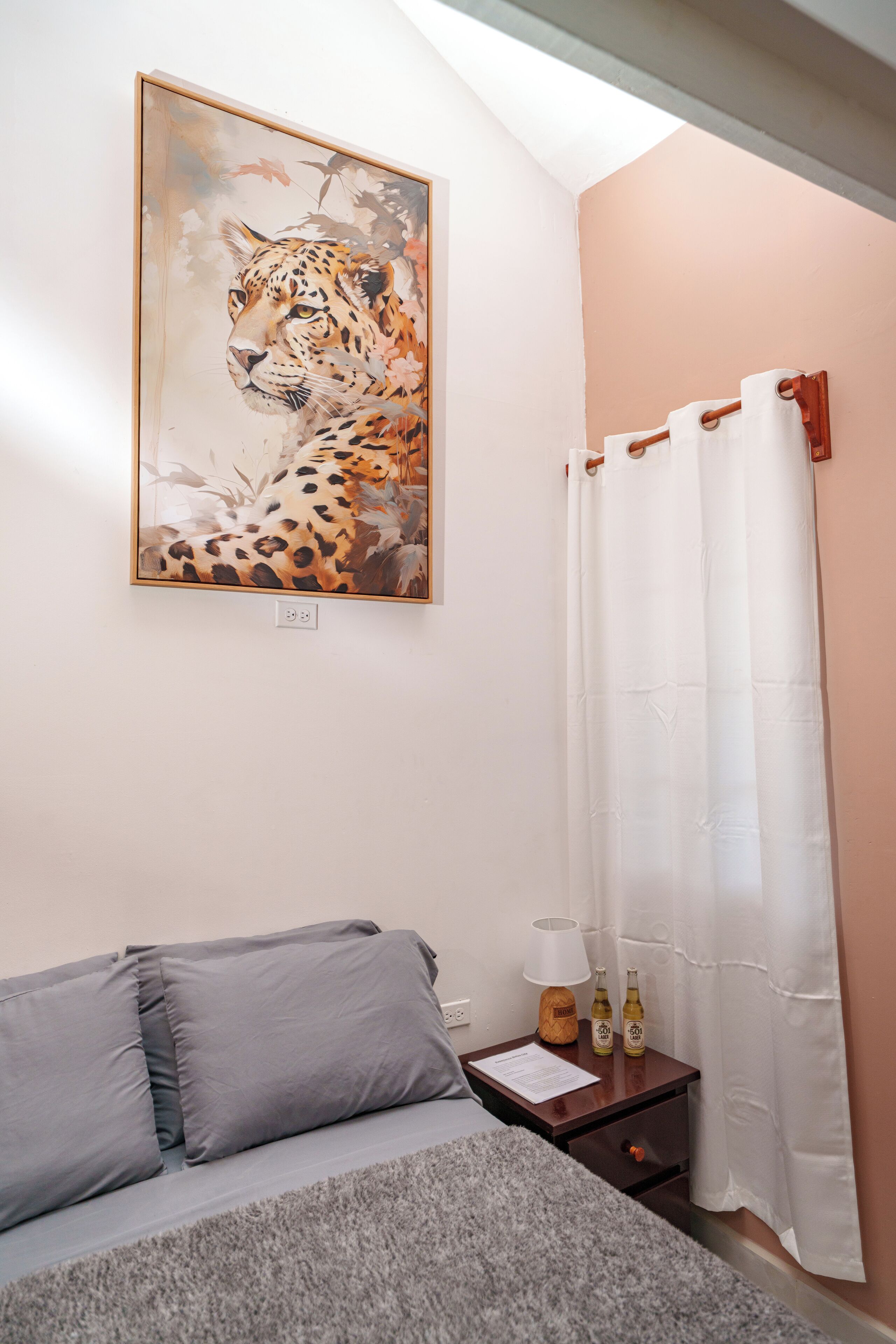 Quarto com cama, travesseiros, mesa de cabeceira com garrafas, abajur e pintura de um leopardo.