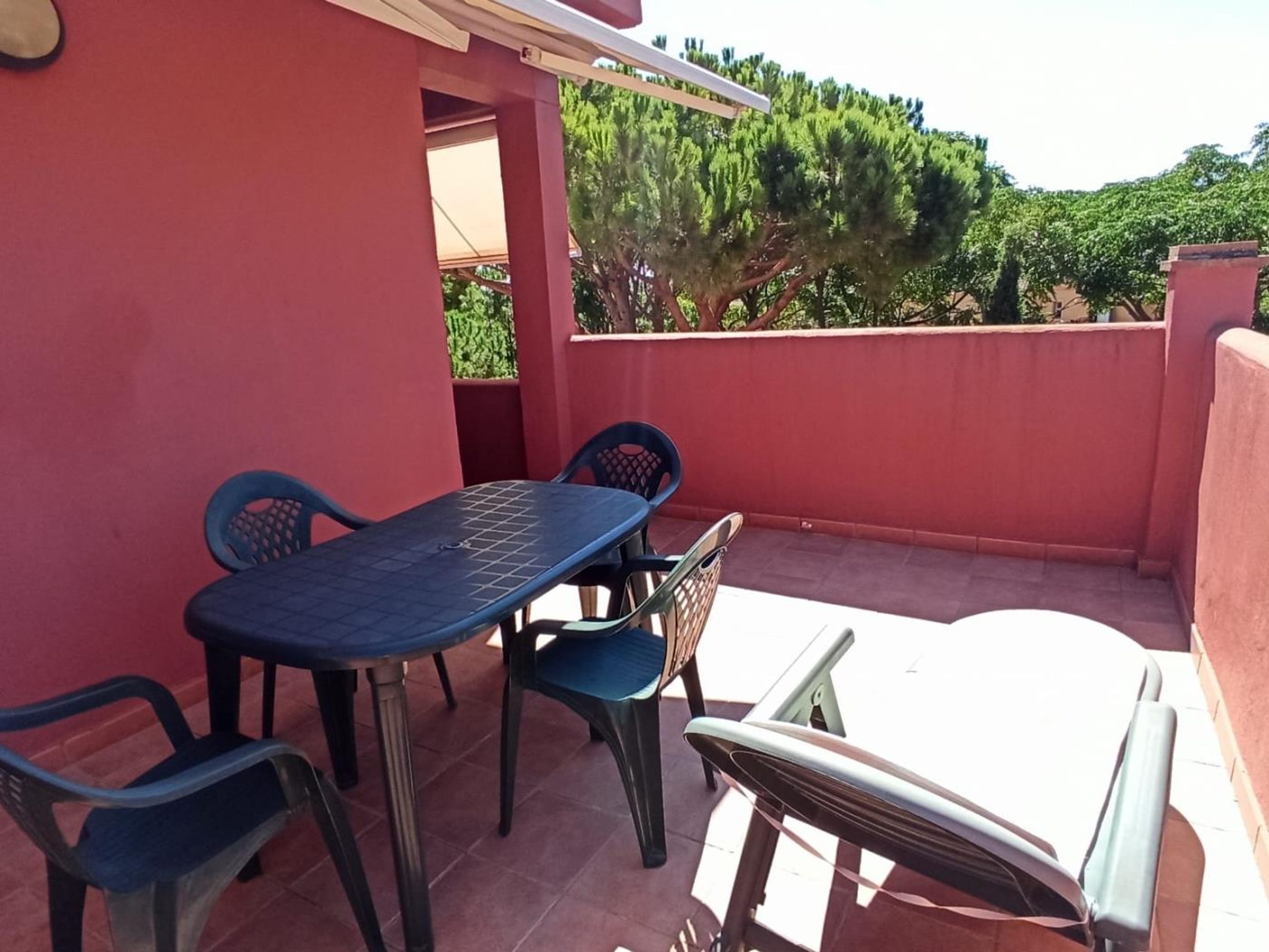 Pequeno pátio ao ar livre com mesa e cadeiras, cercado por um muro vermelho e vista para árvores.