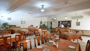 Um restaurante com várias mesas e cadeiras, ventiladores de teto e um balcão com alguém atrás.