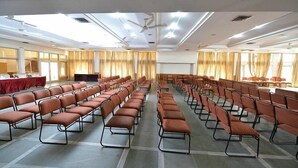 Uma sala de conferências com fileiras de cadeiras, um palco com uma mesa vermelha e amplas janelas com cortinas.