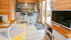 Interior acolhedor de uma cabana com decoração em madeira, uma área de cozinha, uma mesa de jantar com banquetas altas e uma TV sobre um suporte de madeira.