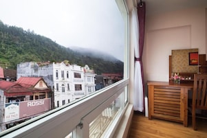 Quarto com vista para uma área montanhosa, uma escrivaninha de madeira e uma janela com cortina.