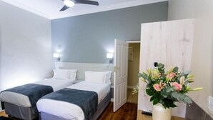 Quarto de hotel com duas camas, um ventilador de teto, piso de madeira e um vaso de flores.