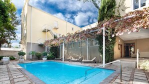 Piscina com deck ladrilhado, área de estar coberta e um edifício com escadaria.