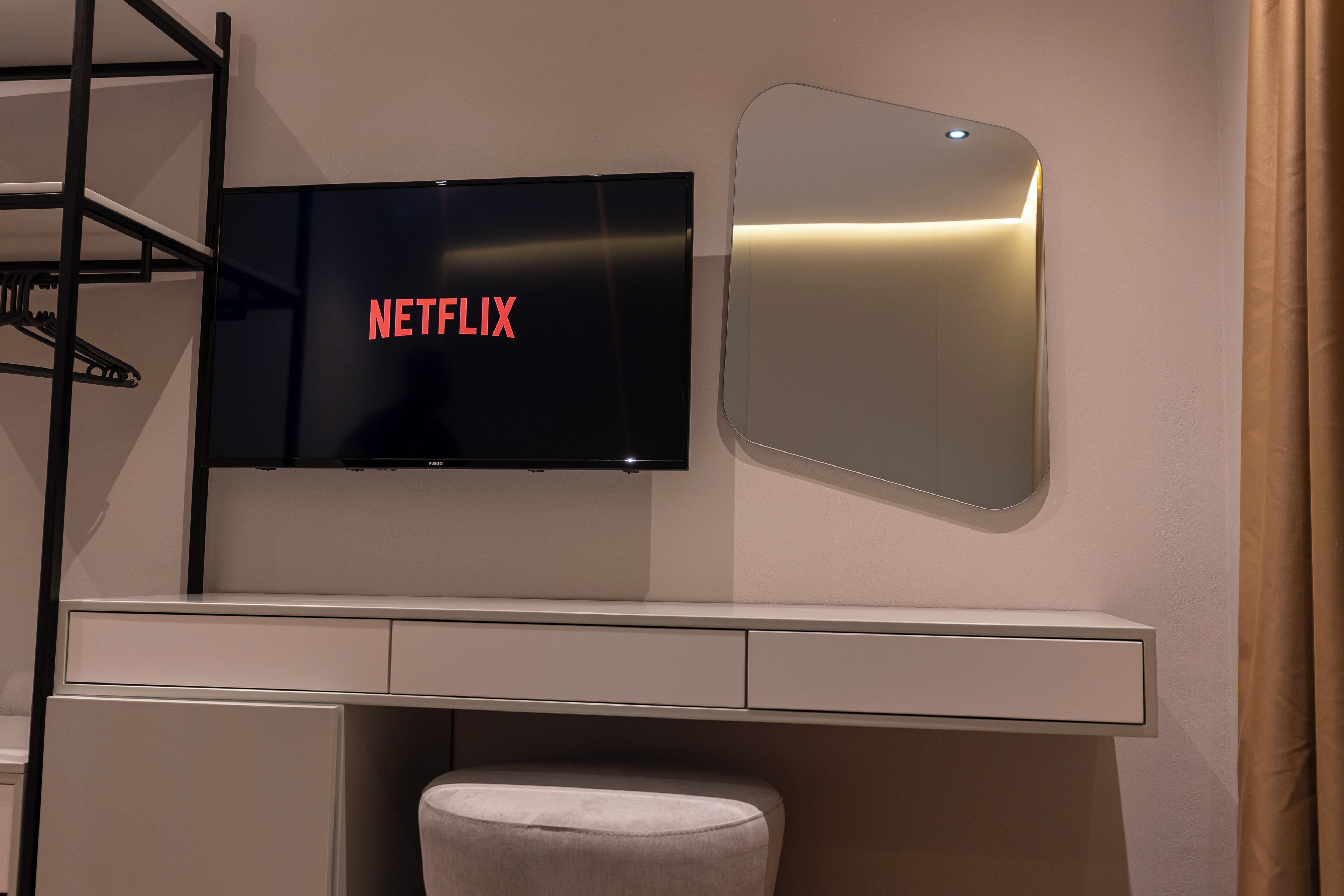 Quarto moderno com TV fixada na parede exibindo 'NETFLIX' em letras vermelhas, uma escrivaninha branca e um pequeno banquinho.