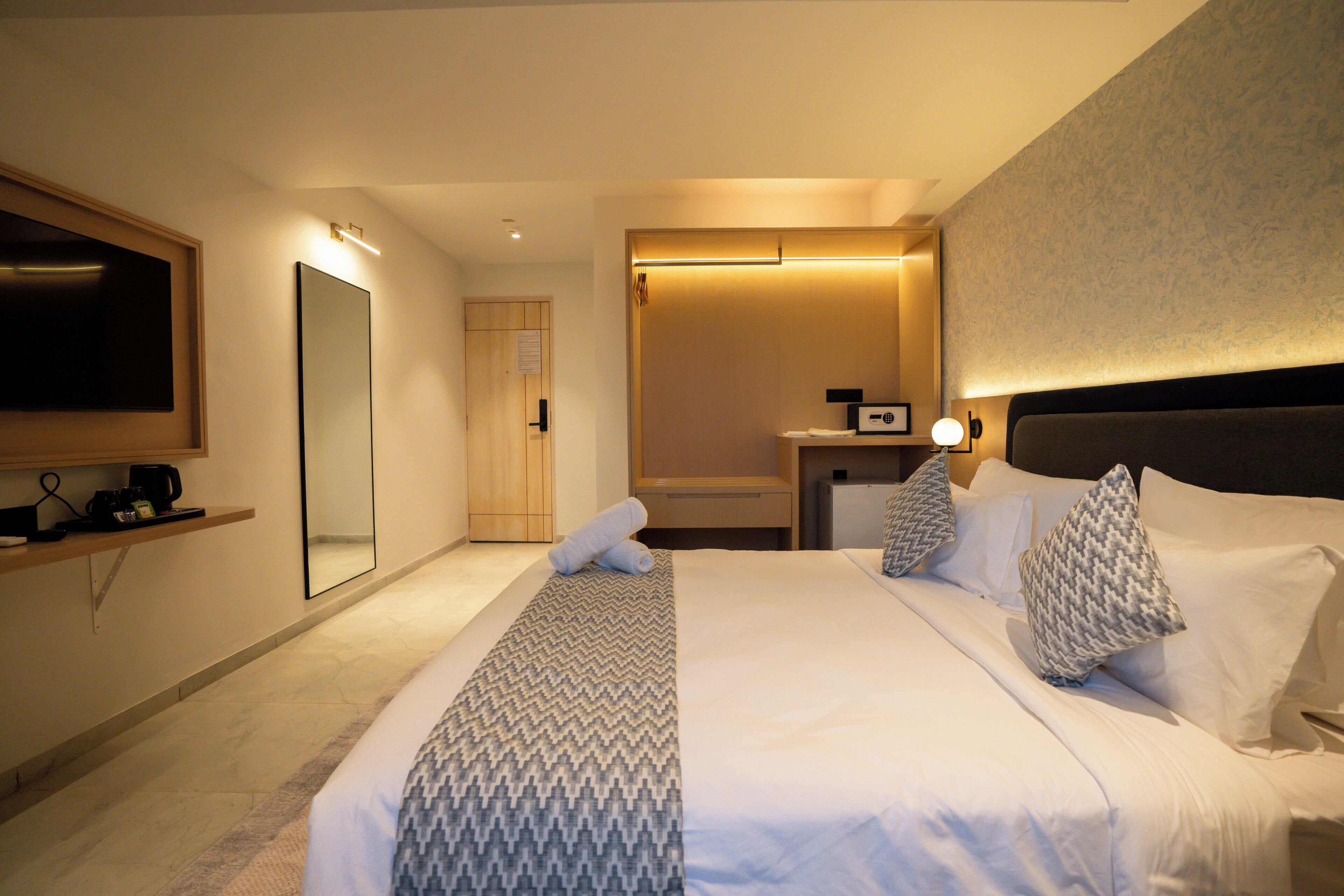 Quarto de hotel com uma cama grande, uma TV de tela plana, um espelho, mesa de cabeceira com abajur e uma pequena escrivaninha com um computador.