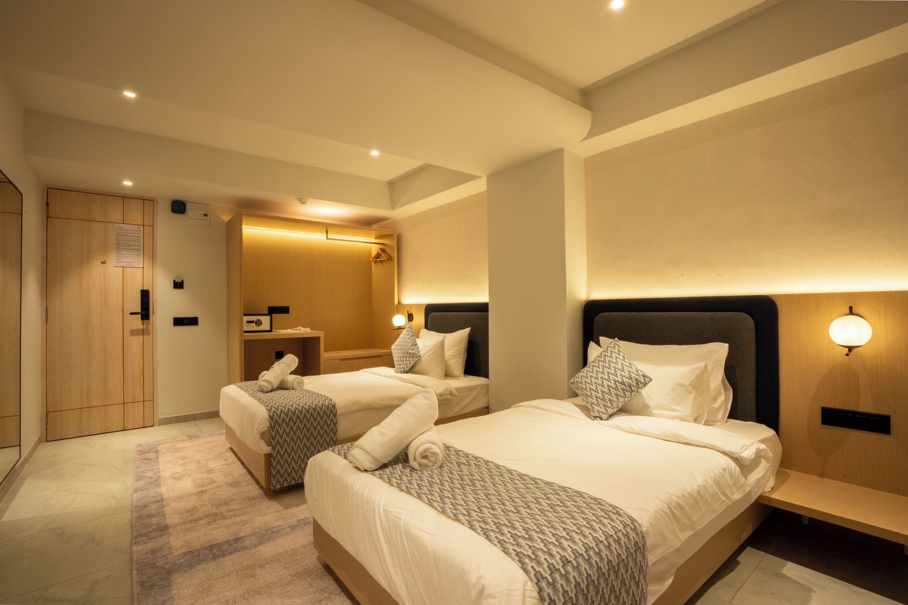 Quarto de hotel moderno com duas camas, um guarda-roupa de madeira, uma mesa de cabeceira e um banco embutido.