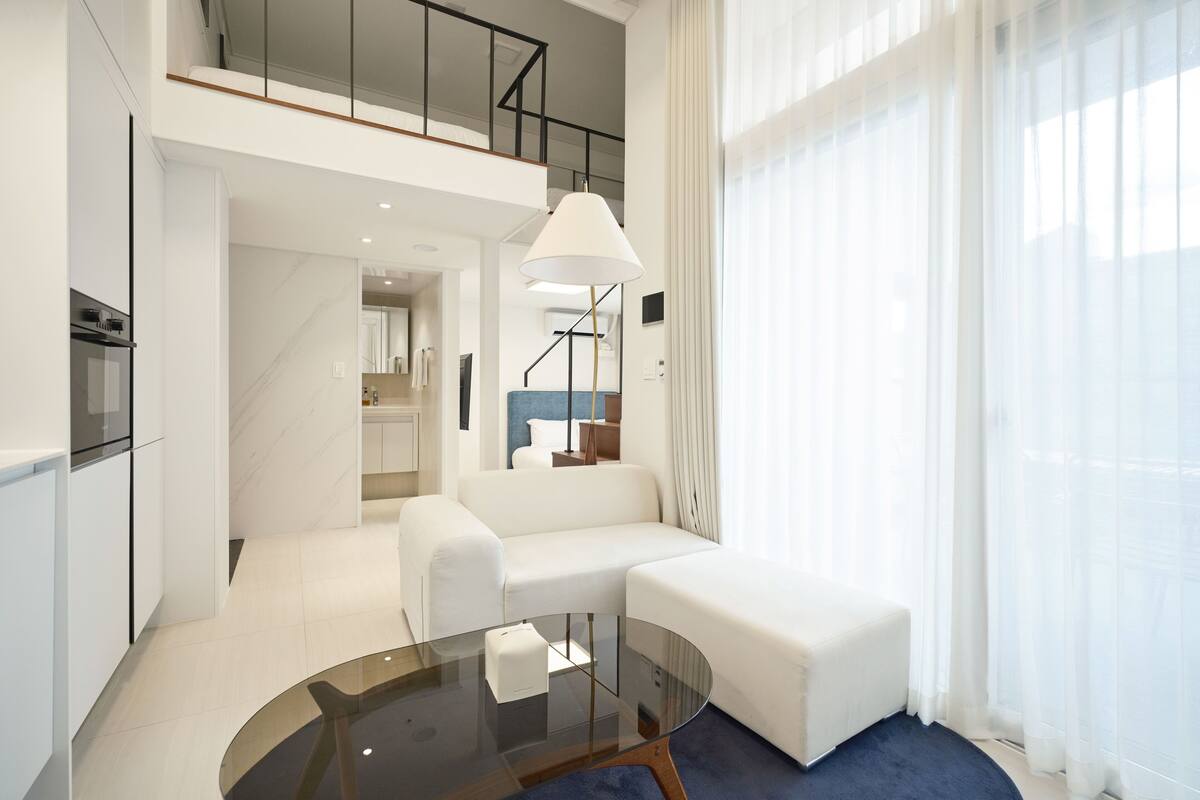 Quarto de hotel moderno com mesa de jantar de vidro, sofá, pequena cozinha e varanda com guarda-corpo.