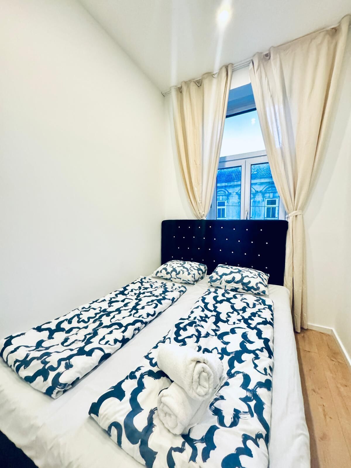 Uma cama de casal com colcha e almofadas estampadas, cabeceira escura e janela com cortinas.