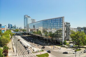 Um edifício moderno com fachada de vidro, uma estrutura de estacionamento em vários níveis e uma rua urbana movimentada com veículos e pedestres.