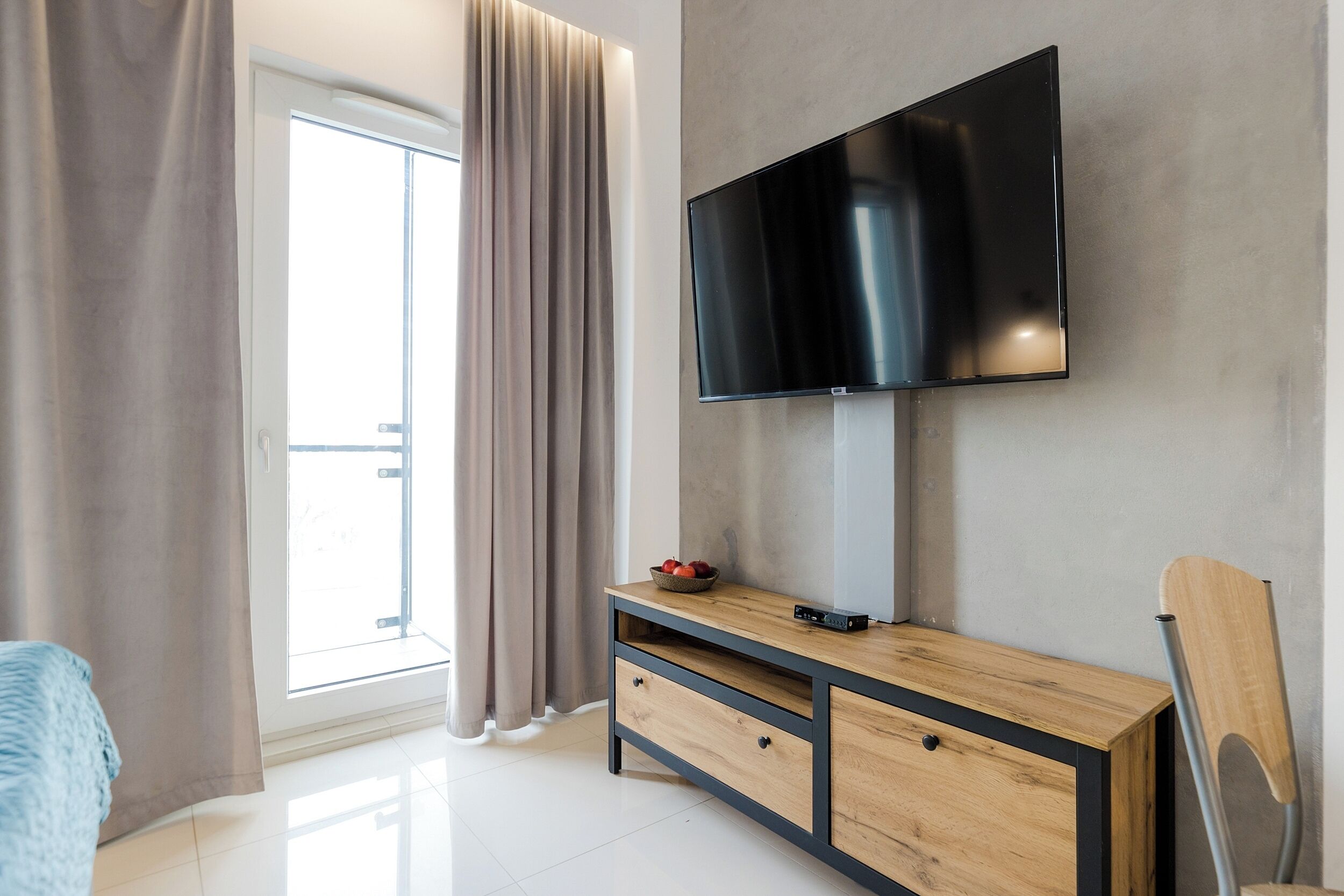 Quarto moderno com televisão fixada na parede, cómoda de madeira, uma cadeira e vista para o exterior por uma janela ampla.