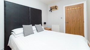 Cama bem arrumada com almofadas brancas e estampadas, cabeceira de madeira, mesa de cabeceira com abajur e porta de madeira com porta-casacos.