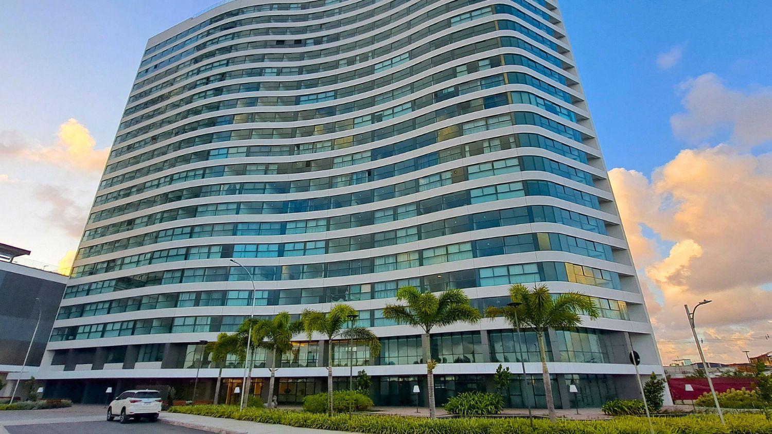 Um edifício moderno de vários andares com fachada curva, grandes janelas e área frontal ajardinada com palmeiras.