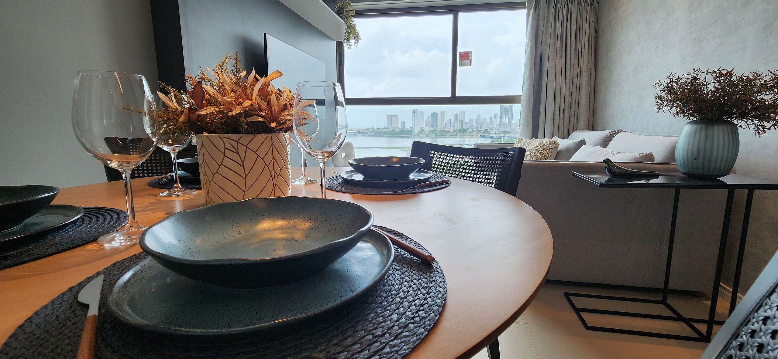 Uma sala de jantar moderna com vista para a cidade, um sofá e uma mesa posta com pratos, tigelas e copos de vinho.