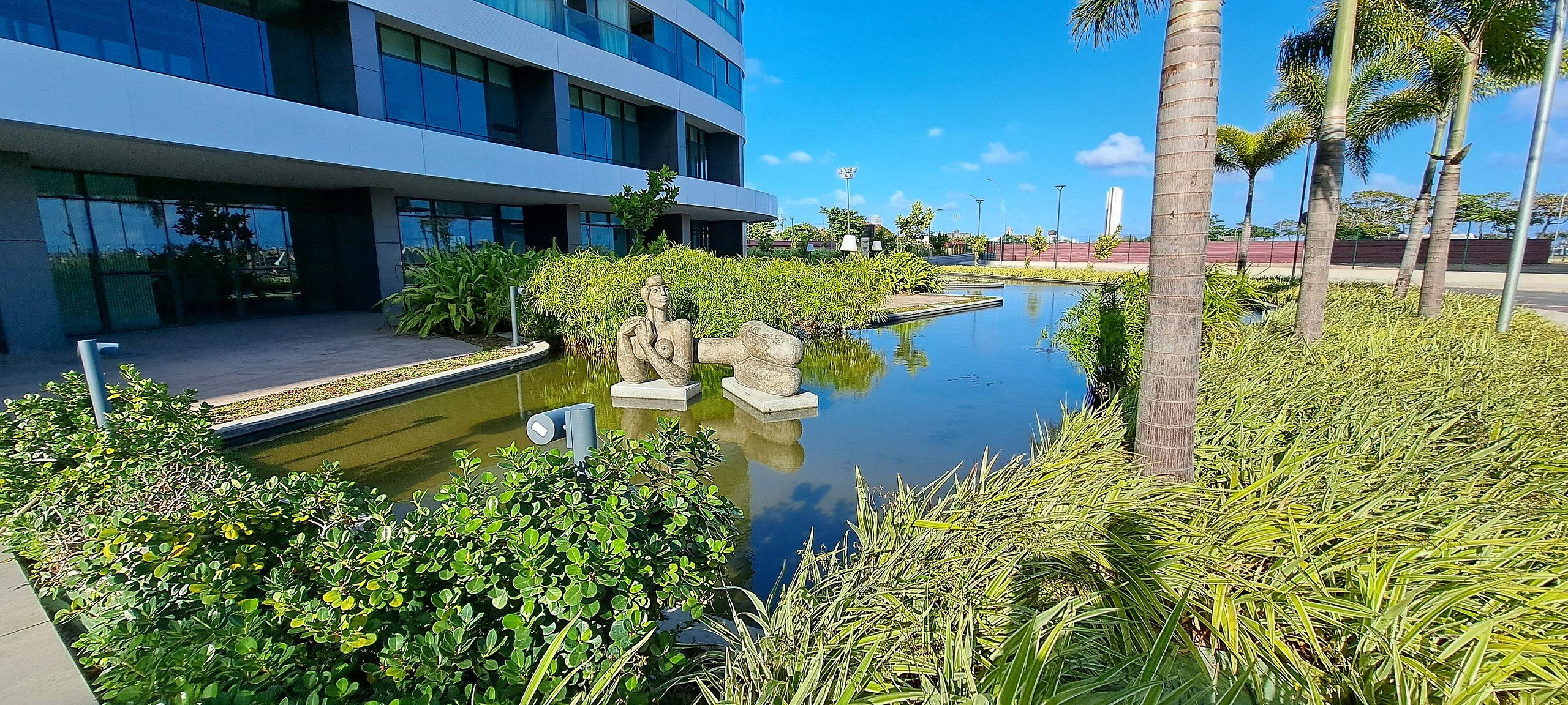 Um edifício moderno com uma piscina refletora, rodeado por vegetação exuberante e palmeiras.