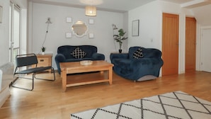 Uma sala de estar moderna com sofá, poltrona e mesa de centro. Há um espelho na parede, uma planta e uma porta ao fundo.