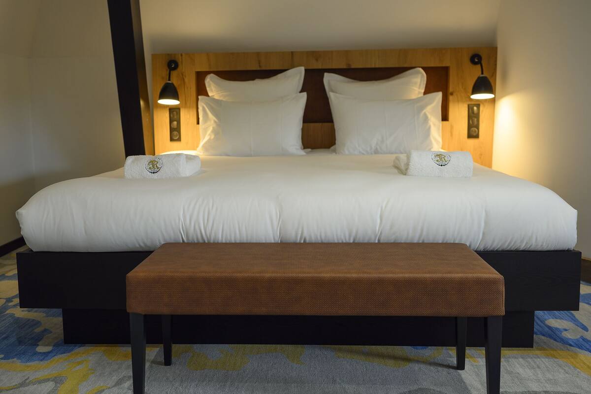 Uma cama grande com roupa de cama branca, duas mesas de cabeceira com abajures e um banco em frente à cama.