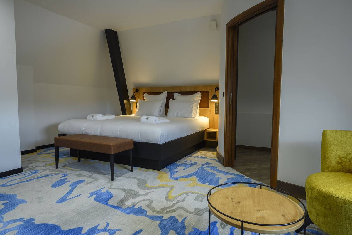 Quarto de hotel com cama, um banco, uma mesa redonda pequena e uma cadeira amarela.