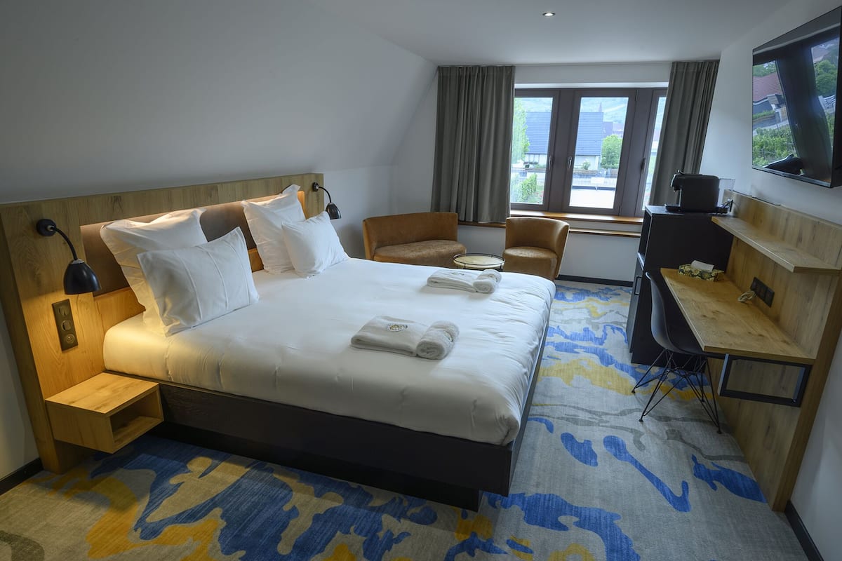 Quarto de hotel com uma cama grande, duas poltronas, uma escrivaninha, televisão de tela plana e janela com cortinas.