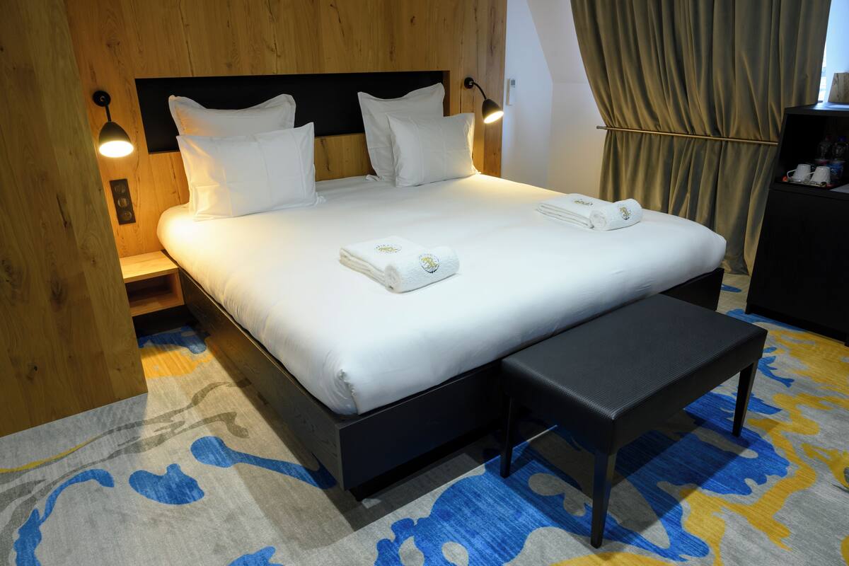 Cama bem arrumada com lençóis brancos e duas toalhas, uma cabeceira de madeira, uma mesa de cabeceira e um banquinho.