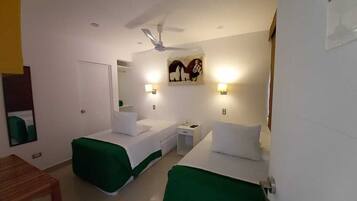Quarto de hotel com duas camas de solteiro, uma escrivaninha, uma cadeira, um ventilador de teto e um quadro na parede.